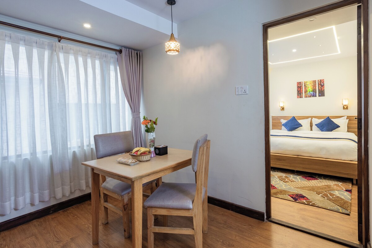 Quarto com mesa de jantar de madeira, duas cadeiras, luminária pendente, cama com colcha estampada e uma obra de arte na parede.