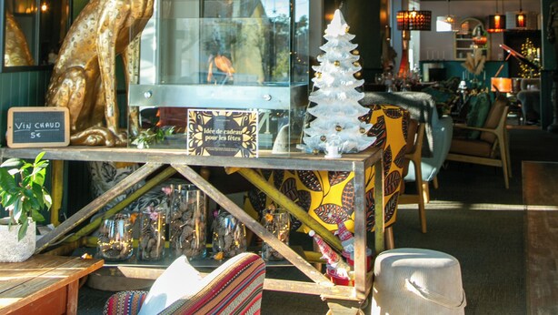 Um interior acolhedor com uma mesa de madeira exibindo vários objetos, uma estátua dourada de gato, um pinheiro de Natal branco e luminárias de vime penduradas.
