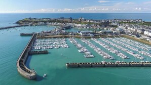 Vista aérea de uma marina com vários barcos atracados, um quebra-mar e uma cidade costeira ao fundo.