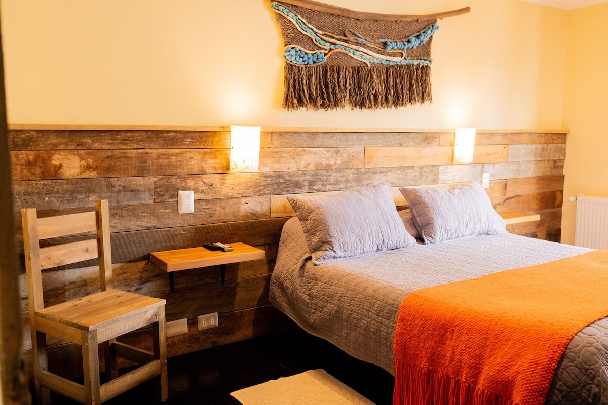 Quarto com cabeceira de madeira, cama com cobertor cinza, cadeira de madeira, mesa de cabeceira com abajur e um objeto decorativo na parede.