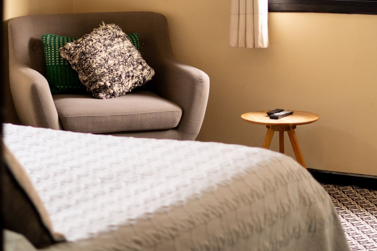 Um quarto com uma cama, uma cadeira, uma mesinha e uma janela com cortinas.