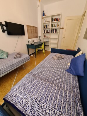 Quarto com duas camas, uma escrivaninha, uma cadeira, uma estante e uma televisão.