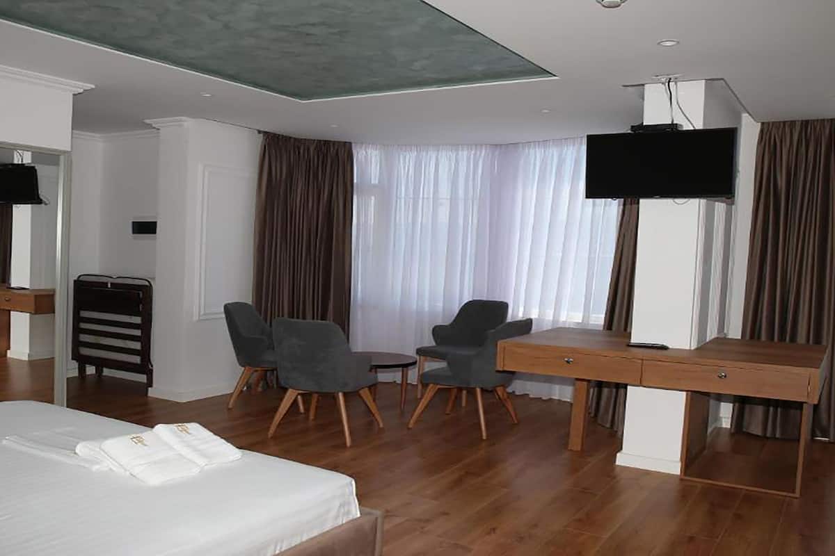 Quarto de hotel com cama, escrivaninha, duas cadeiras, televisão e janela com cortinas.