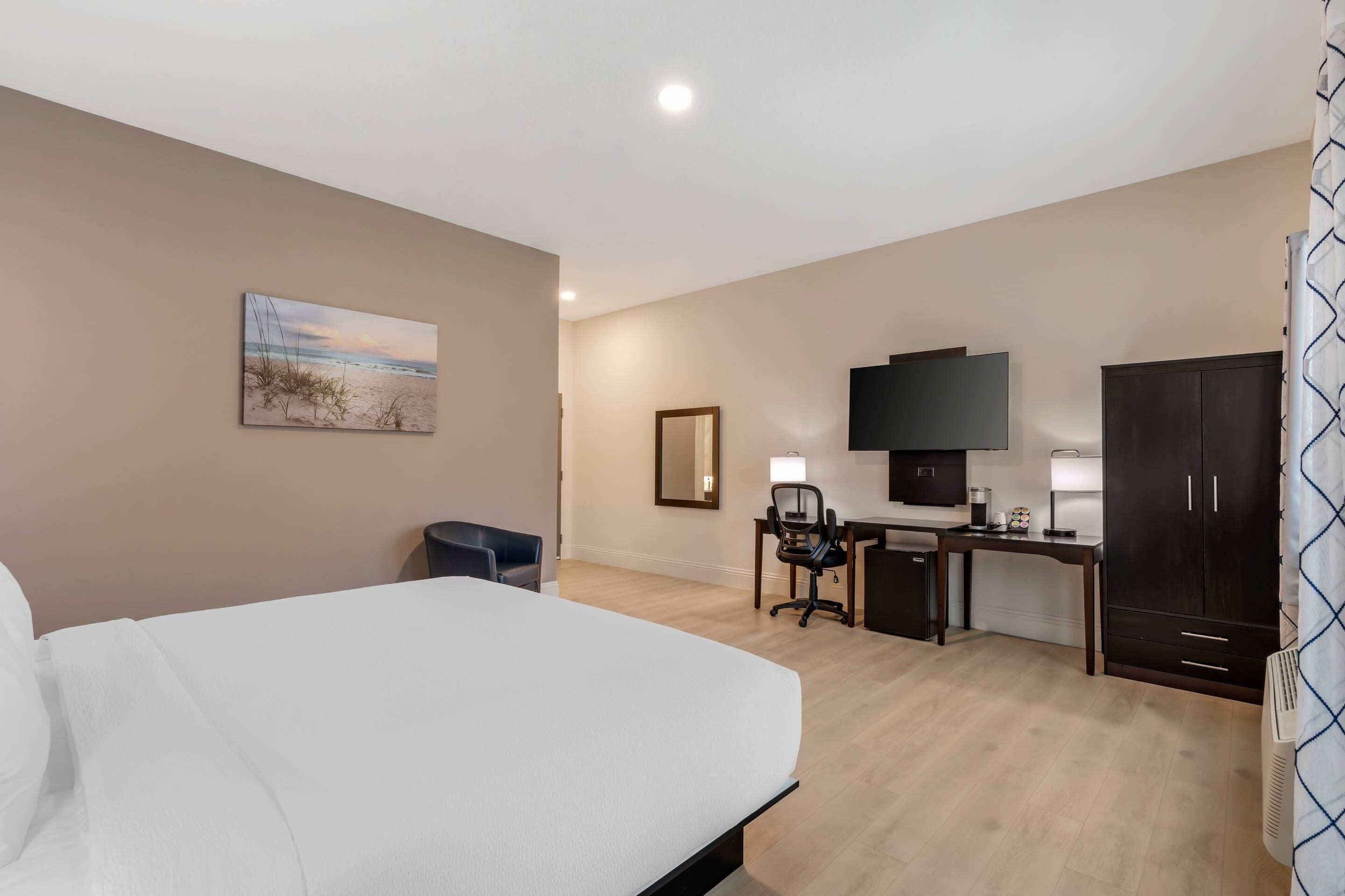 Quarto de hotel com cama, escrivaninha, cadeira, televisão, guarda-roupa e um quadro na parede.