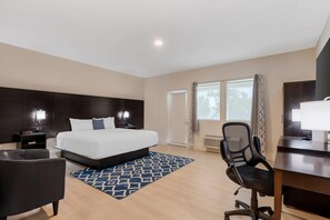 Quarto de hotel com uma cama grande, uma escrivaninha, uma cadeira, uma mesinha de cabeceira e uma televisão.