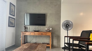 Uma sala de estar moderna com uma mesa de centro de madeira, uma TV fixada numa parede cinzenta, uma pequena mesa lateral de madeira com objetos decorativos e um ventilador de pé.