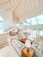 Uma sala de estar ampla, com teto alto, escada que leva aos andares superiores e grandes janelas com vista para o exterior.