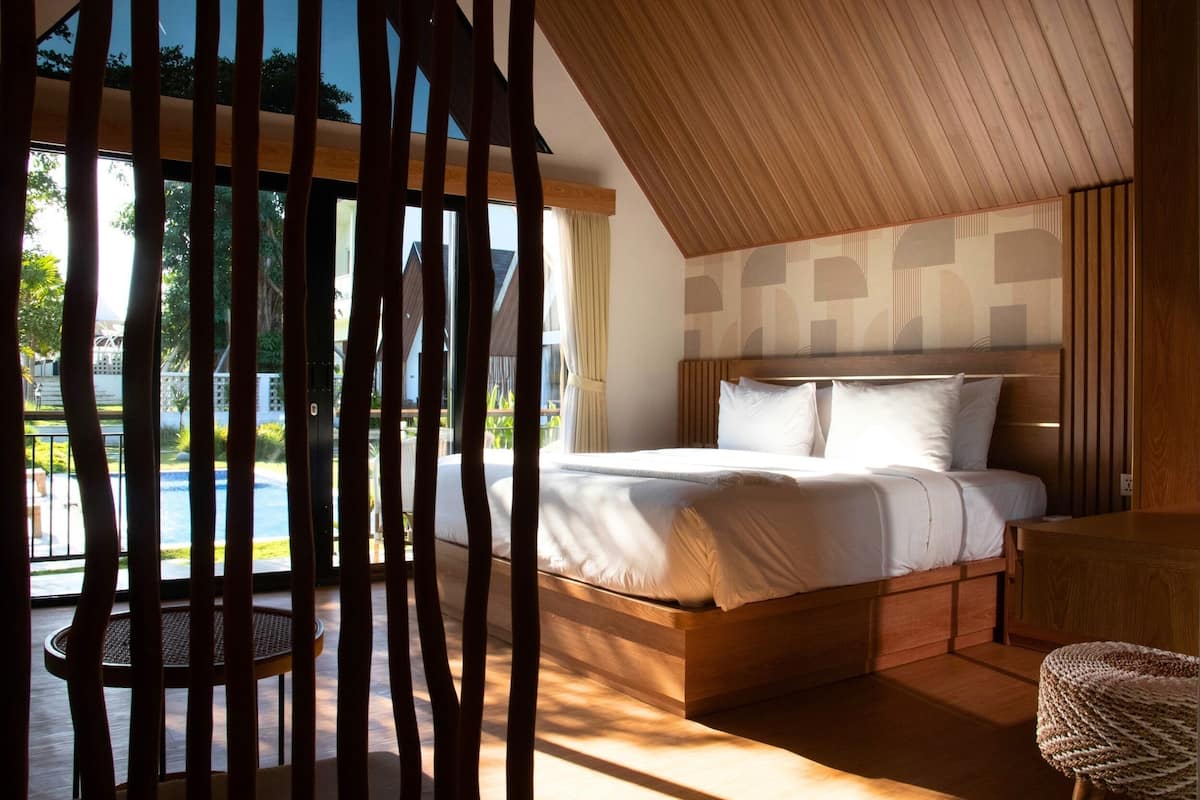 Quarto com cama, cabeceira de madeira, janela com cortinas e vista para uma piscina.