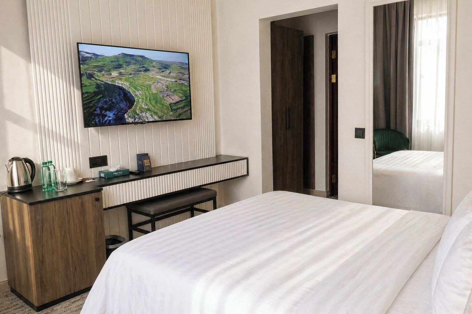 Quarto de hotel com uma cama, uma escrivaninha com televisão de tela plana, um banco e uma porta que dá para outro quarto.