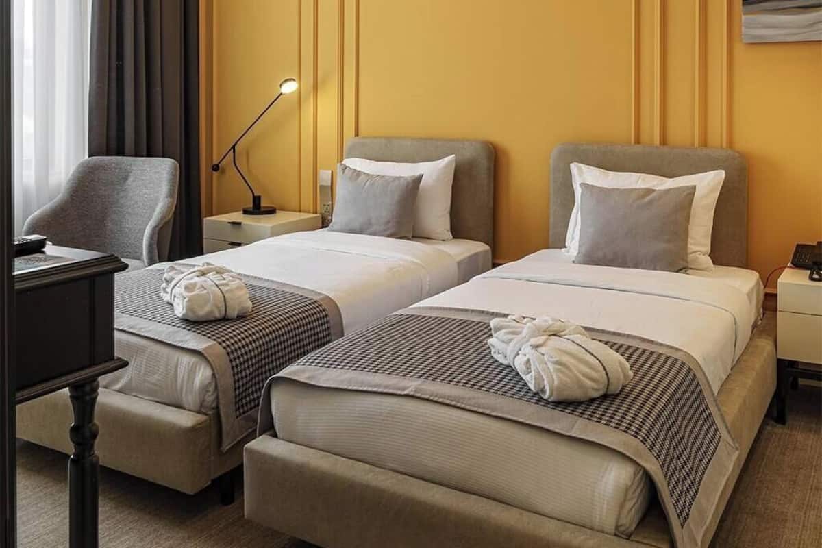 Quarto de hotel com duas camas, uma mesa, uma cadeira, uma luminária e um quadro na parede.