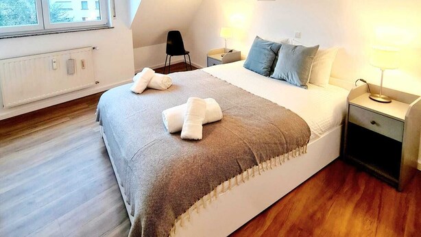 Quarto moderno com cama grande, piso de madeira, radiador, janela e luminária pendente.