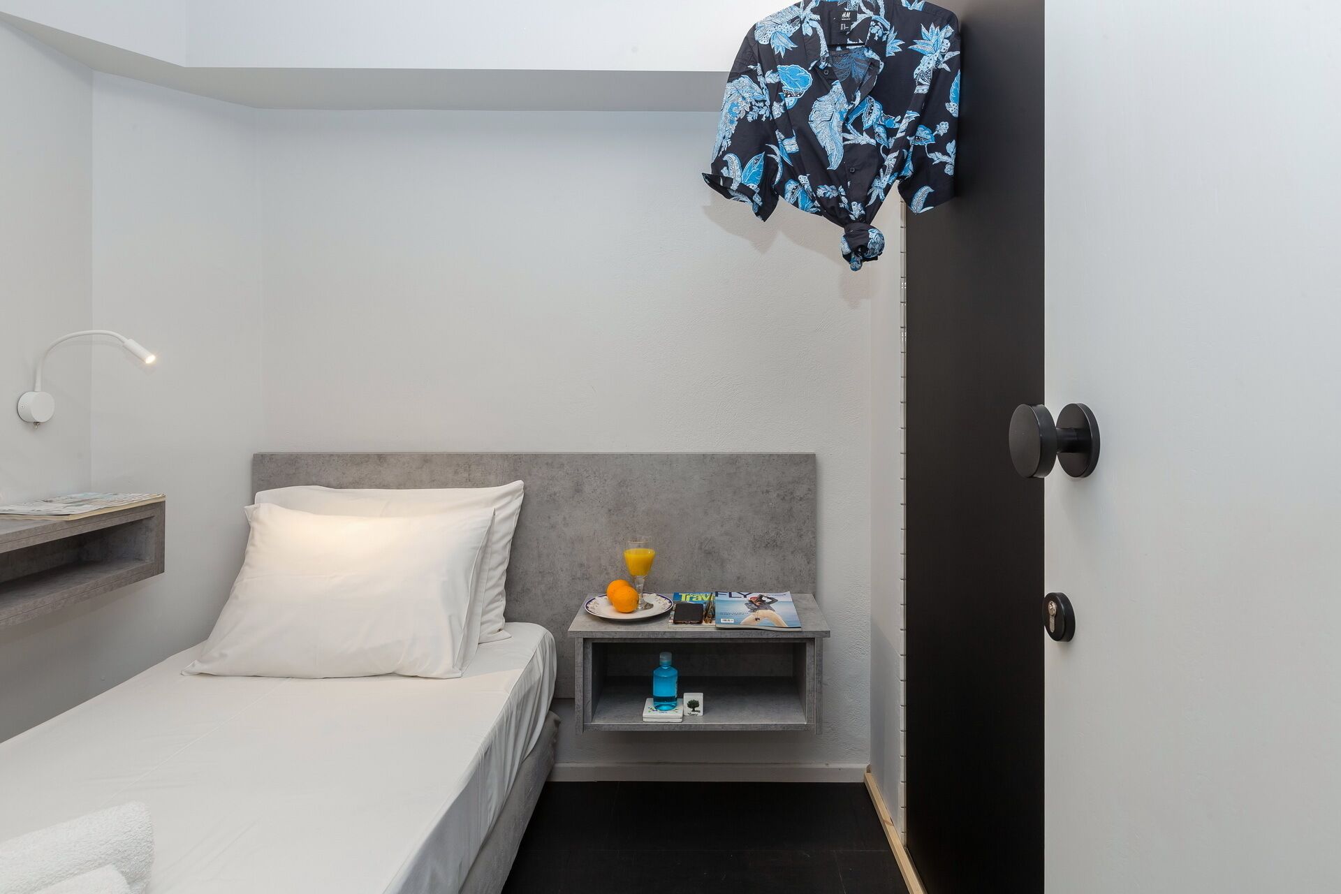 Quarto de hotel moderno com cama, mesa de cabeceira com luminária, criado-mudo com bebida e revista, e porta com puxador.