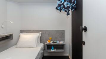 Quarto de hotel moderno com cama, mesa de cabeceira com luminária, criado-mudo com bebida e revista, e porta com puxador.