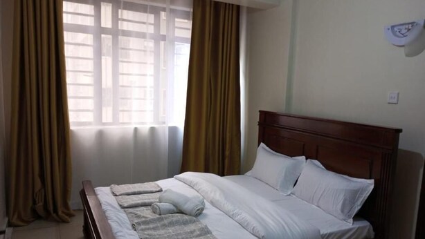 Cama bem arrumada com roupa de cama branca e cabeceira de madeira em quarto com janela e cortinas.