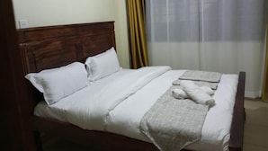 Cama bem arrumada com lençóis brancos e manta cinza, cabeceira de madeira e janela com cortinas leves.