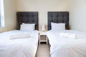 Quarto de hotel com duas camas, cada uma com roupa de cama branca e toalha dobrada.