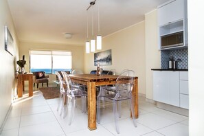 Uma cozinha moderna com armários brancos, uma mesa de jantar de madeira com cadeiras transparentes e uma pintura da paisagem urbana na parede.