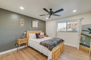 Quarto com cama de madeira, ventoinha no teto, mesa de cabeceira e um criado-mudo com abajur, além de janela com cortinas.