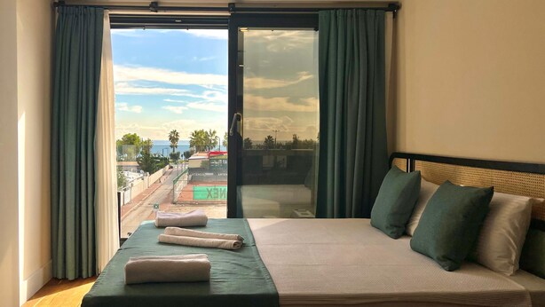 Quarto de hotel com cama, almofadas e vista para a rua e palmeiras.
