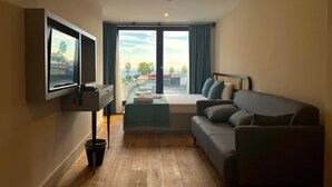 Quarto de hotel compacto com cama, sofá, televisão e janela com vista para palmeiras e edifícios.