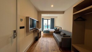Quarto de hotel moderno com piso de madeira, sofá cinza, cama com cabeceira azul, televisão montada na parede e varanda com cortinas.