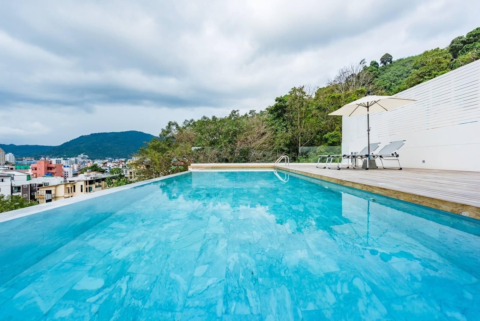 Uma piscina azul cristalina com um deck de madeira, cadeiras de descanso e uma parede branca com uma porta.