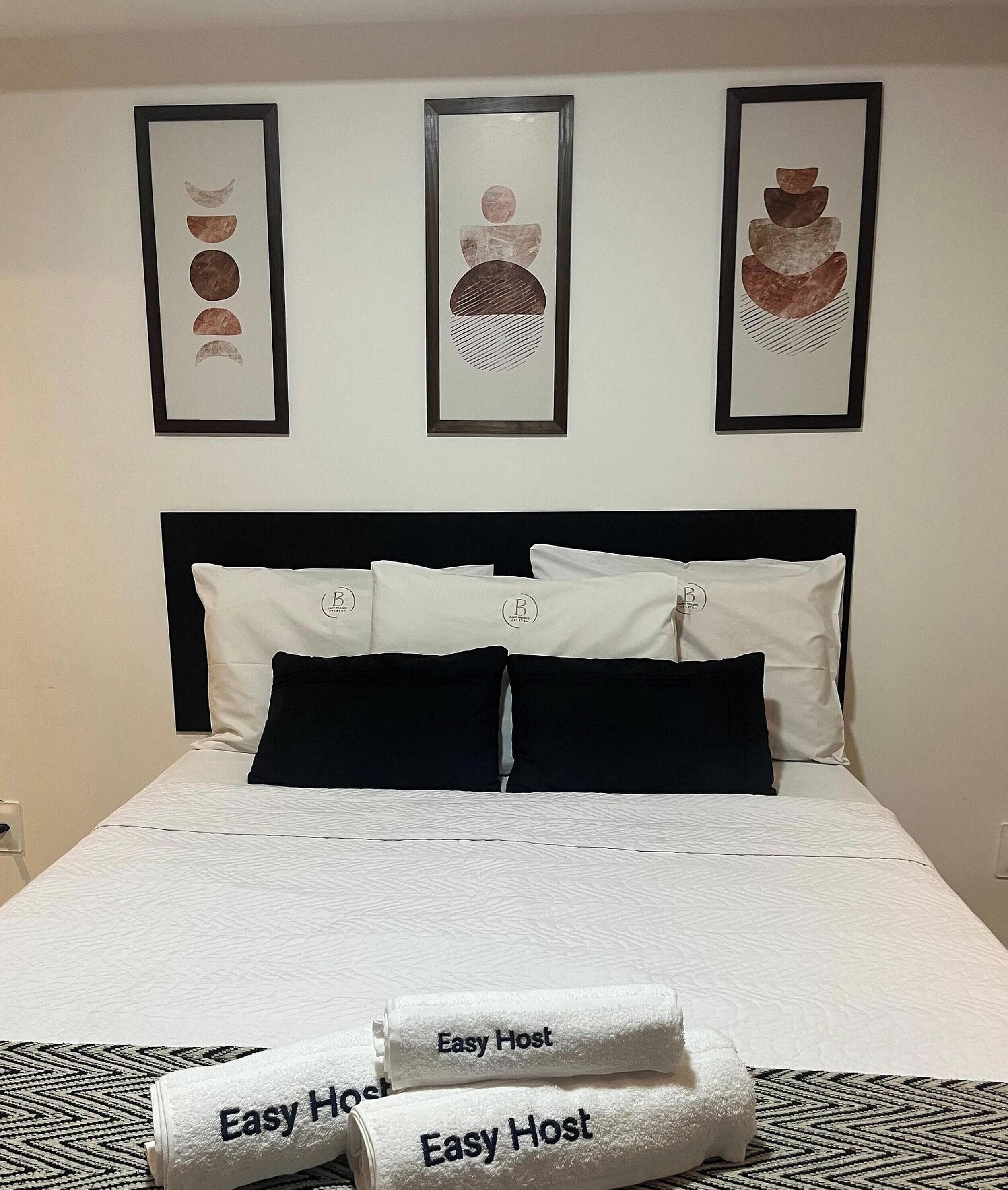 Uma cama bem arrumada com roupa de cama branca e duas almofadas, uma cabeceira e três quadros na parede acima.