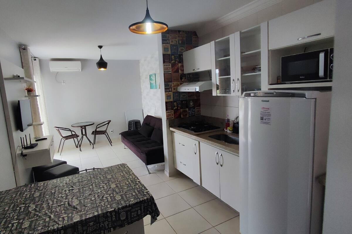 Cozinha compacta com armários brancos, micro-ondas e geladeira. Possui área de refeições com mesa e cadeiras, e um sofá.