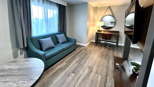Quarto de hotel moderno com sofá verde-água, piso de madeira, uma mesa redonda e uma escrivaninha com abajur.