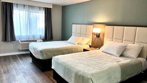 Quarto de hotel com duas camas, uma janela grande com cortinas, ar-condicionado fixado na parede e uma mesa de cabeceira com abajur.