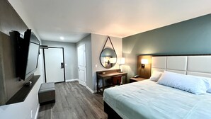 Quarto de hotel com uma cama, uma escrivaninha com cadeira, televisão, espelho e mesa de cabeceira com abajur.