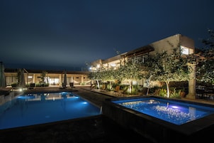 Vista noturna de uma área de piscina de hotel, com duas piscinas, espreguiçadeiras e um edifício com janelas iluminadas.