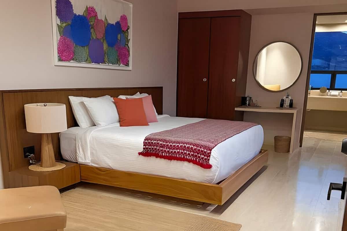 Quarto de hotel moderno com uma cama grande, uma mesinha de cabeceira com luminária, um banco de madeira, um tapete e um banheiro com espelho e pia.