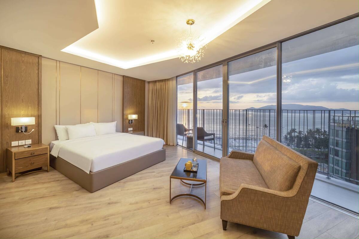 Quarto de hotel com uma cama grande, um sofá, uma mesa pequena e uma varanda com vista para a cidade.