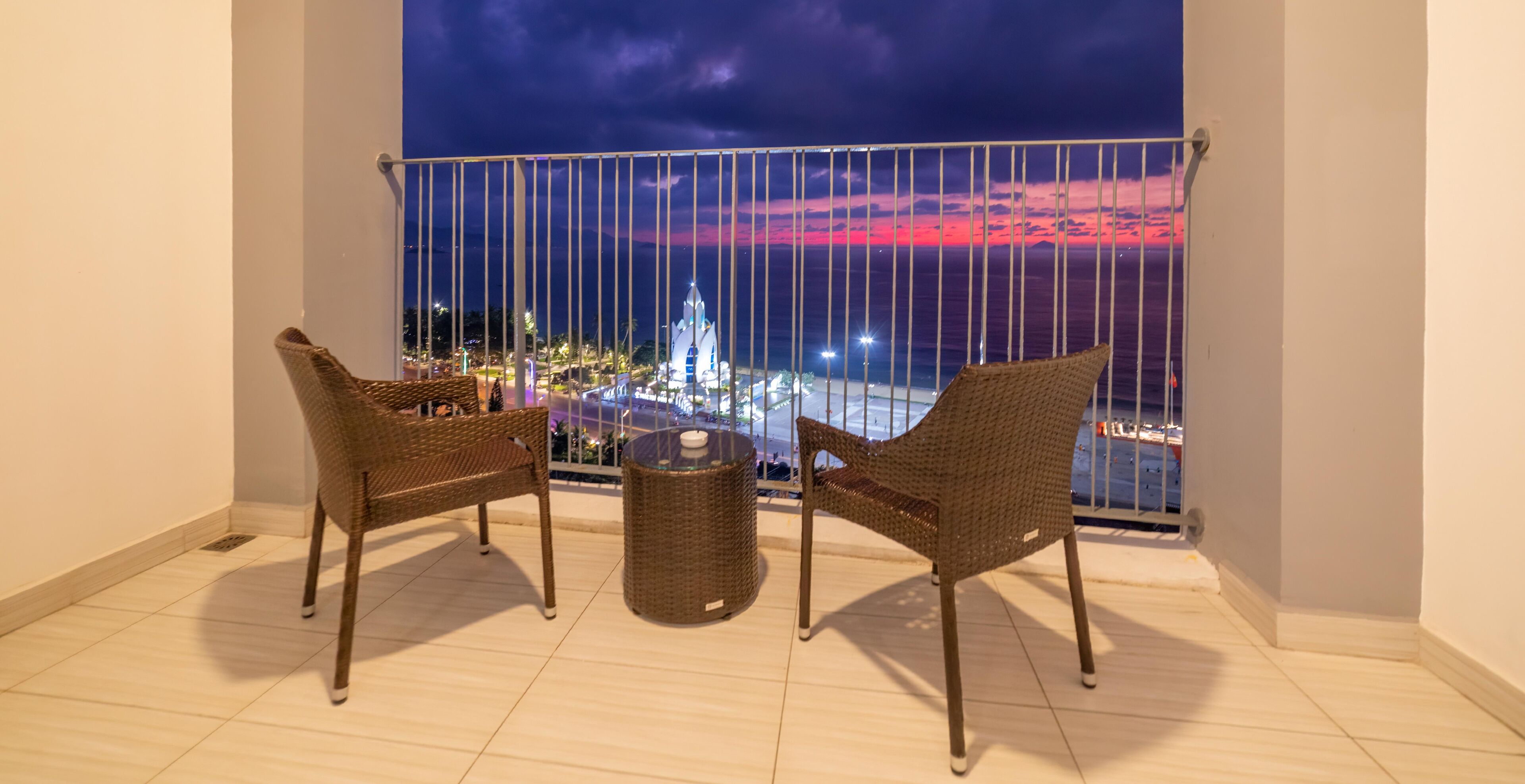 Varanda com cadeiras de vime e uma pequena mesa, com vista para a paisagem urbana, destacando um edifício e um céu colorido ao entardecer.