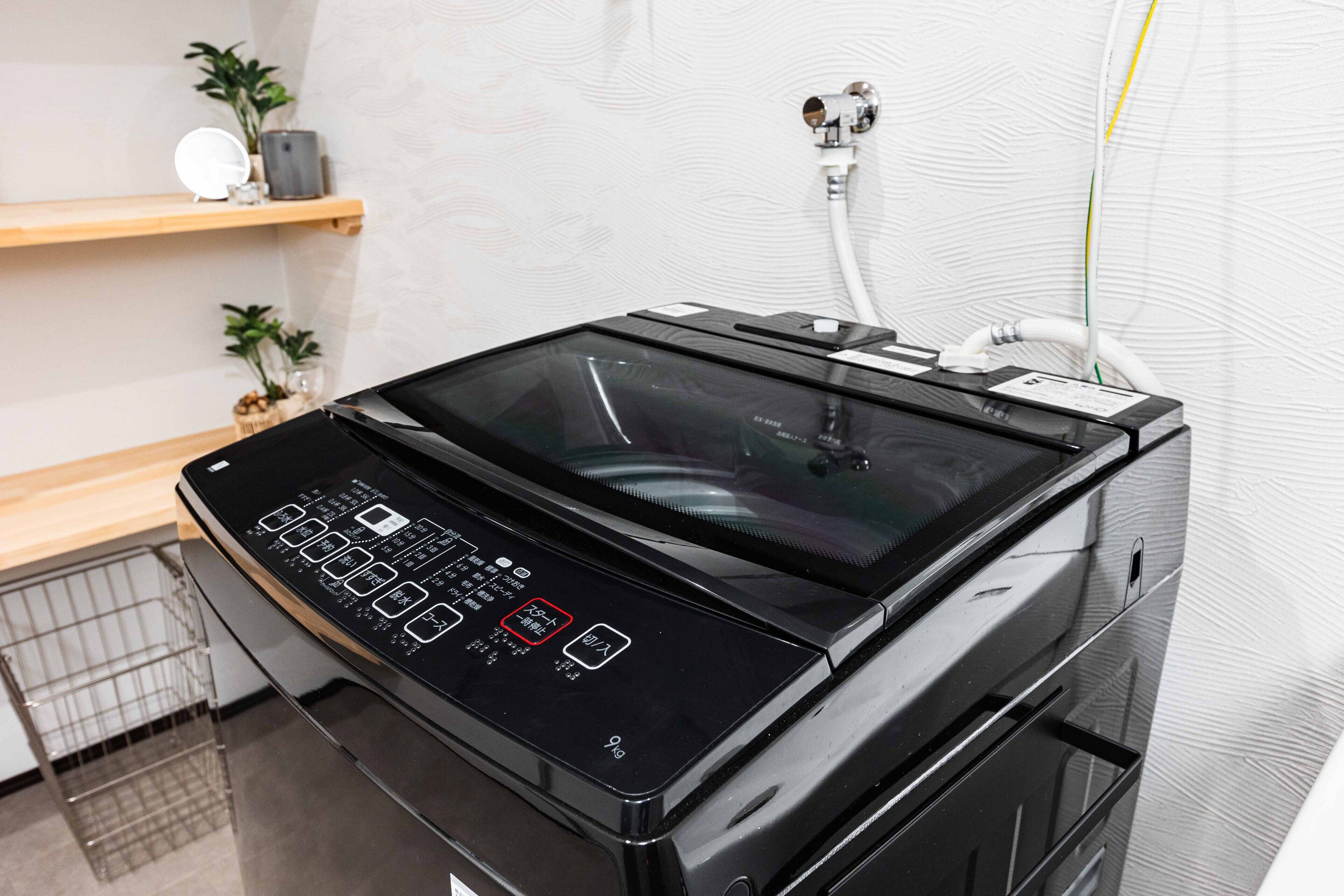 Máquina de lavar roupa preta, compacta e moderna, fixada na parede, com painel de comandos, num espaço limpo e minimalista com prateleiras de madeira e uma pequena planta.