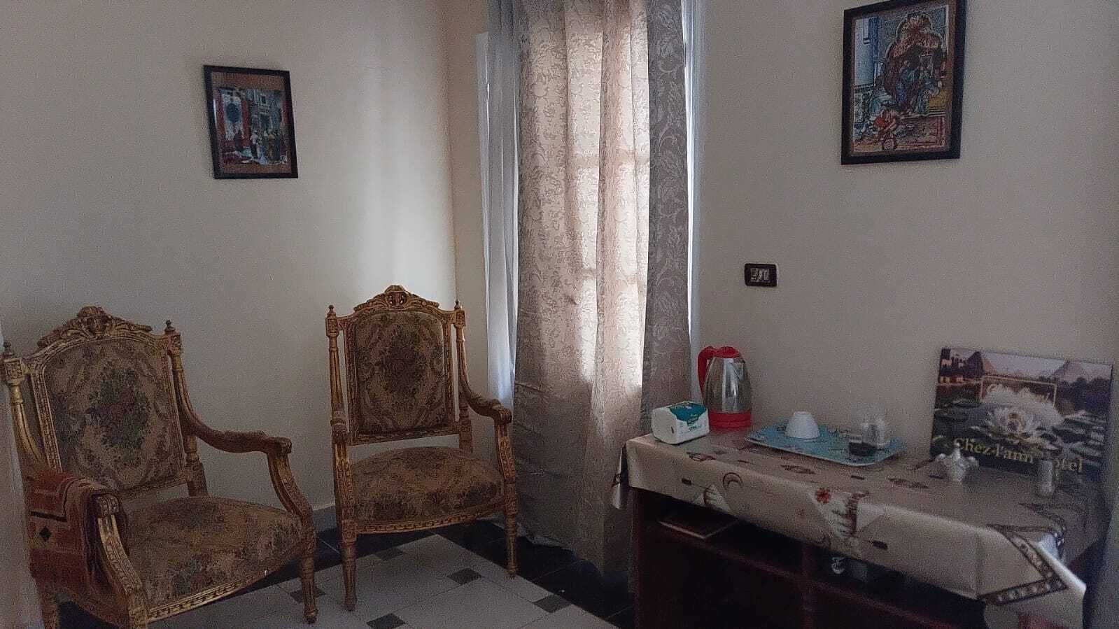 Quarto com duas cadeiras de madeira ornamentadas, uma mesa com toalha, chaleira e um quadro emoldurado na parede.