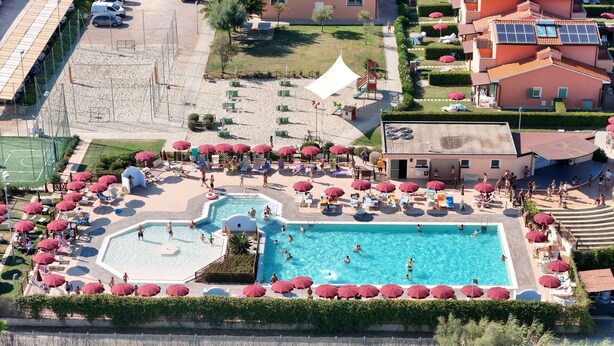 Uma grande piscina ao ar livre cercada por guarda-sóis vermelhos, com uma quadra de tênis e um estacionamento visíveis ao fundo.