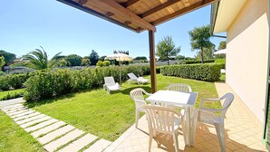 Uma área de pátio com mesa e cadeiras, uma pérgula e um jardim bem cuidado, com cerca viva e palmeira.