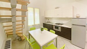 Cozinha moderna com armários brancos, fogão a gás e geladeira. Há uma mesa de jantar com cadeiras verdes e uma pequena planta sobre ela. Uma escada em espiral é visível no lado esquerdo da imagem.