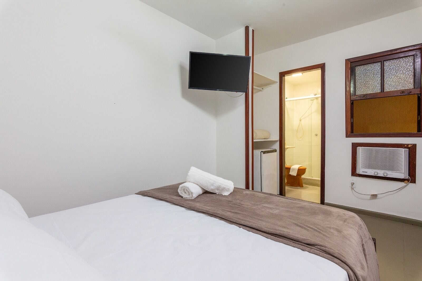 Quarto de hotel com cama, televisão fixada na parede, área de chuveiro visível através de uma porta de vidro e um alto-falante na parede.