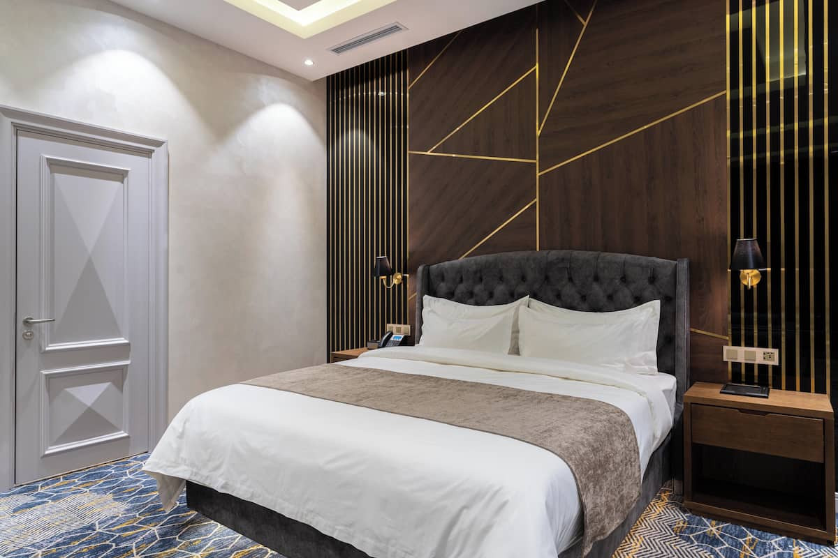 Quarto de hotel moderno com uma cama grande, cabeceira de madeira, mesa de cabeceira com abajur e tapete estampado.