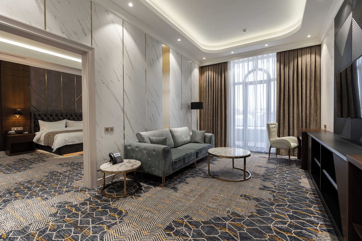 Quarto de hotel com cama, sofá, duas mesas de cabeceira, televisão e uma janela grande com cortinas.