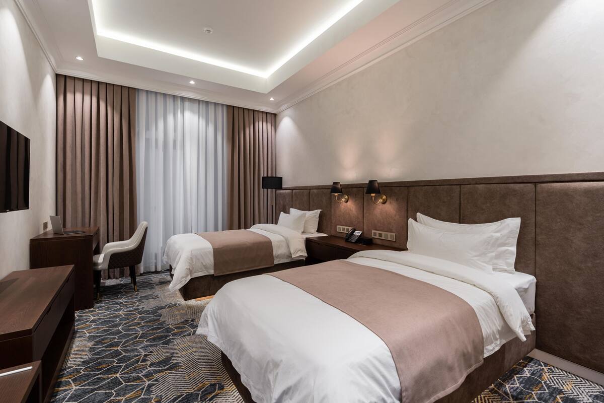 Quarto de hotel com duas camas, uma mesa, uma cadeira e uma janela grande com cortinas.