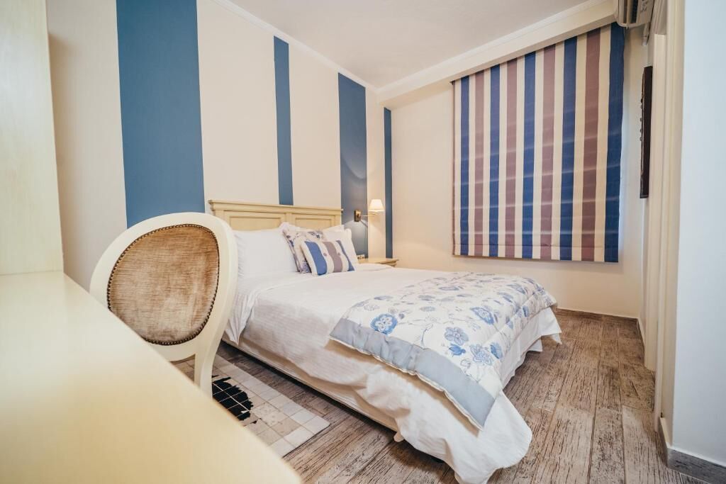 Quarto de hotel com cama, cadeira, mesa de cabeceira e janela com cortinas listradas.