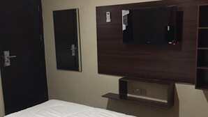 Quarto de hotel com cama, porta escura, televisão na parede e uma pequena prateleira.