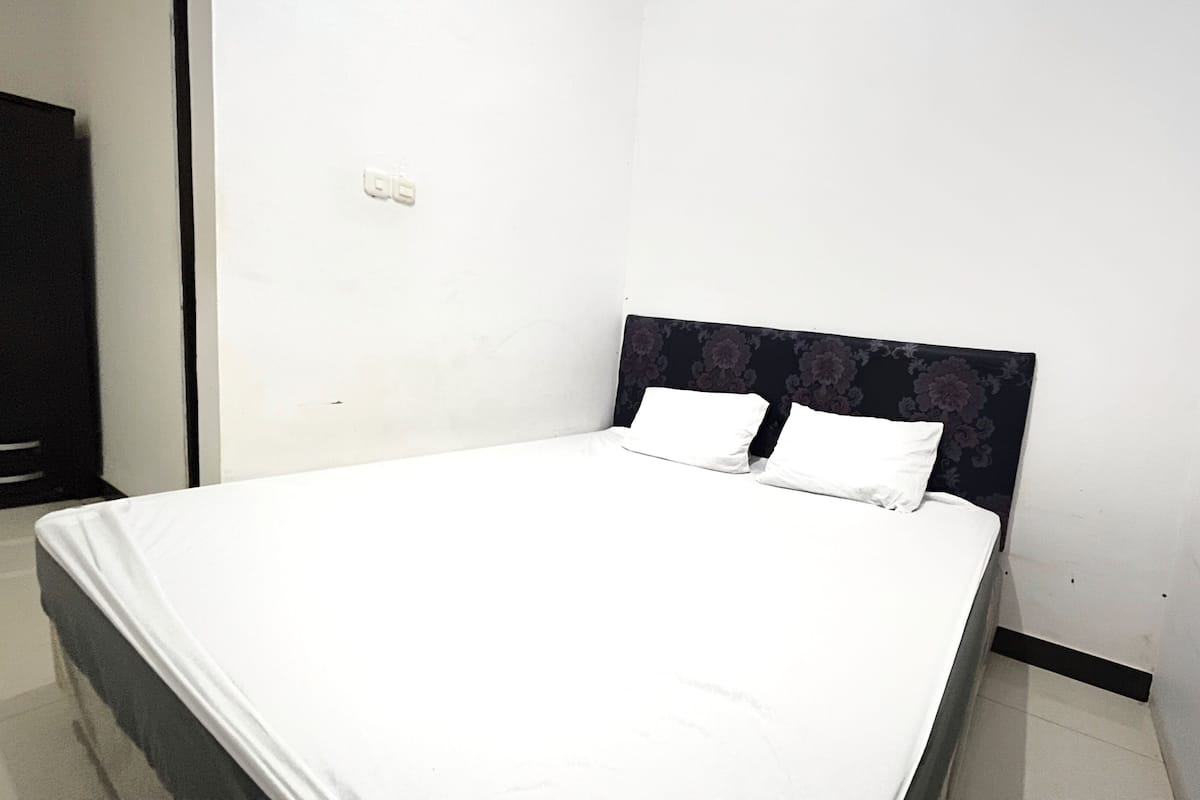 Uma cama de solteiro com cabeceira escura e duas almofadas brancas em quarto com paredes brancas e chão preto.