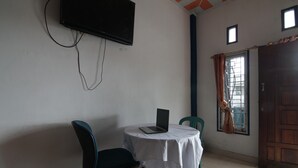 Um quarto pequeno com uma mesa redonda, uma cadeira, um laptop, uma TV fixada na parede, uma janela com cortinas e uma porta de madeira.