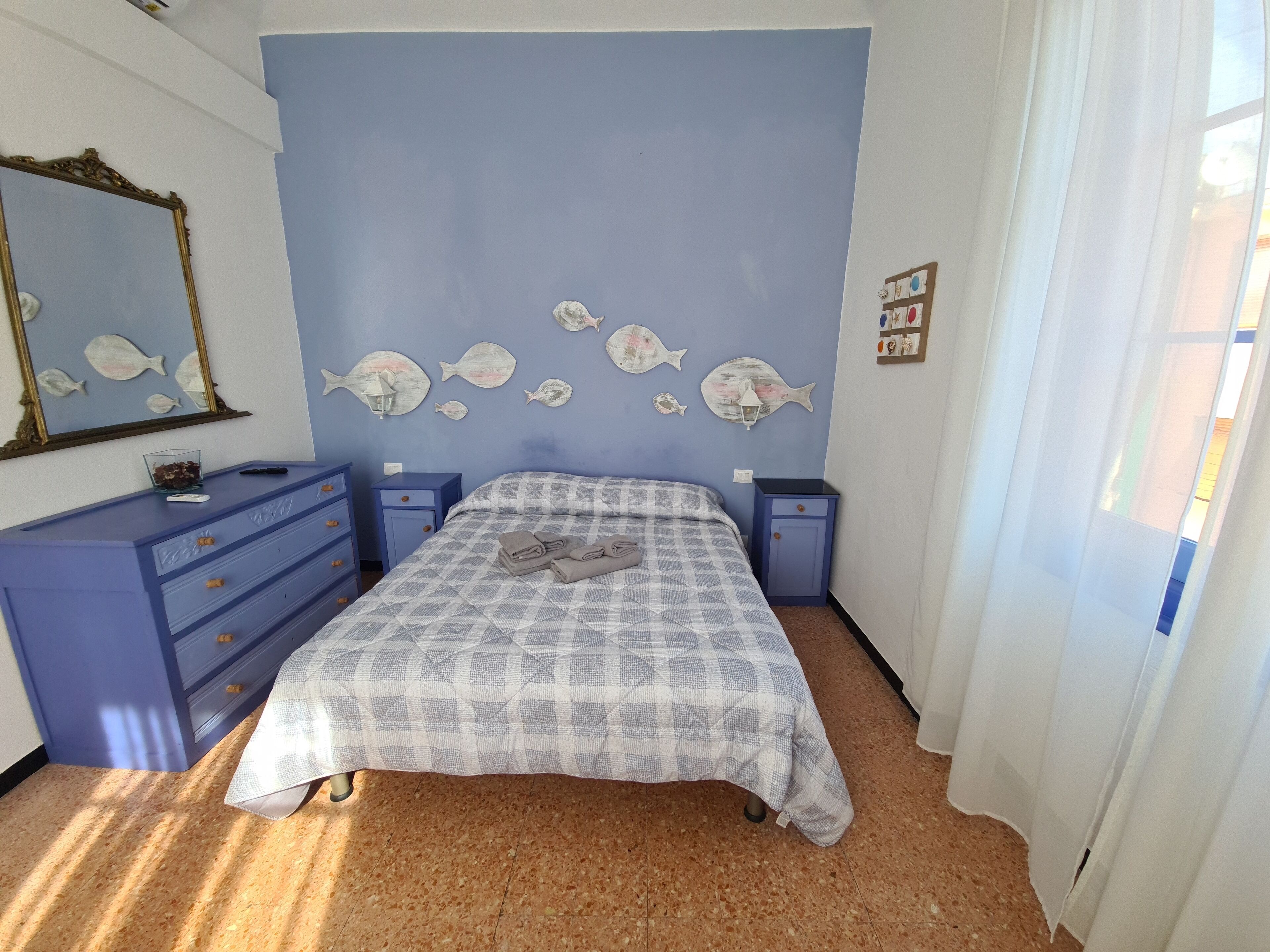 Quarto com parede azul, cama com colcha xadrez, cômoda, mesa de cabeceira e arte na parede com desenhos de peixes.