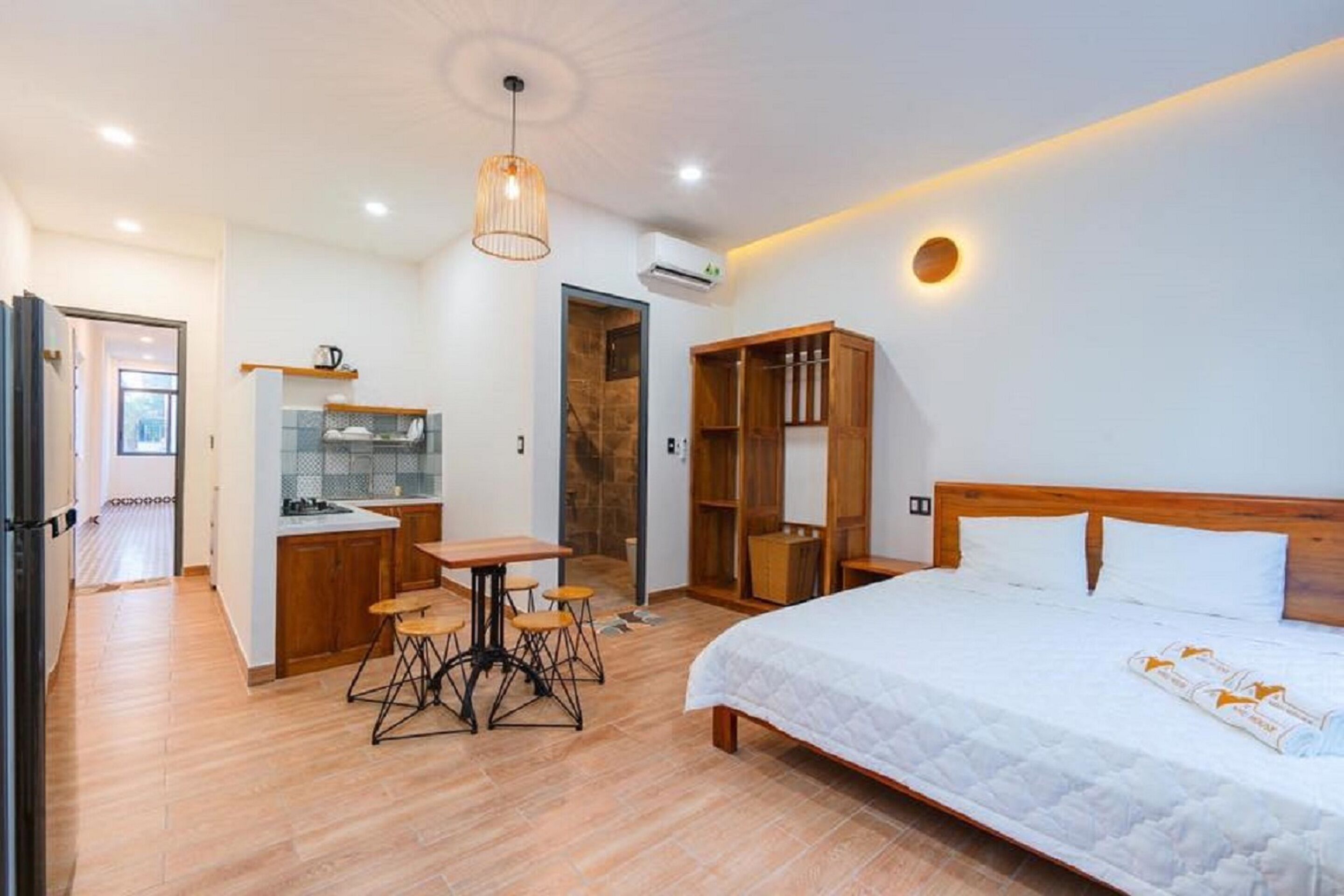 Quarto com cama, guarda-roupa de madeira, kitchenette e área de refeições com bancos.
